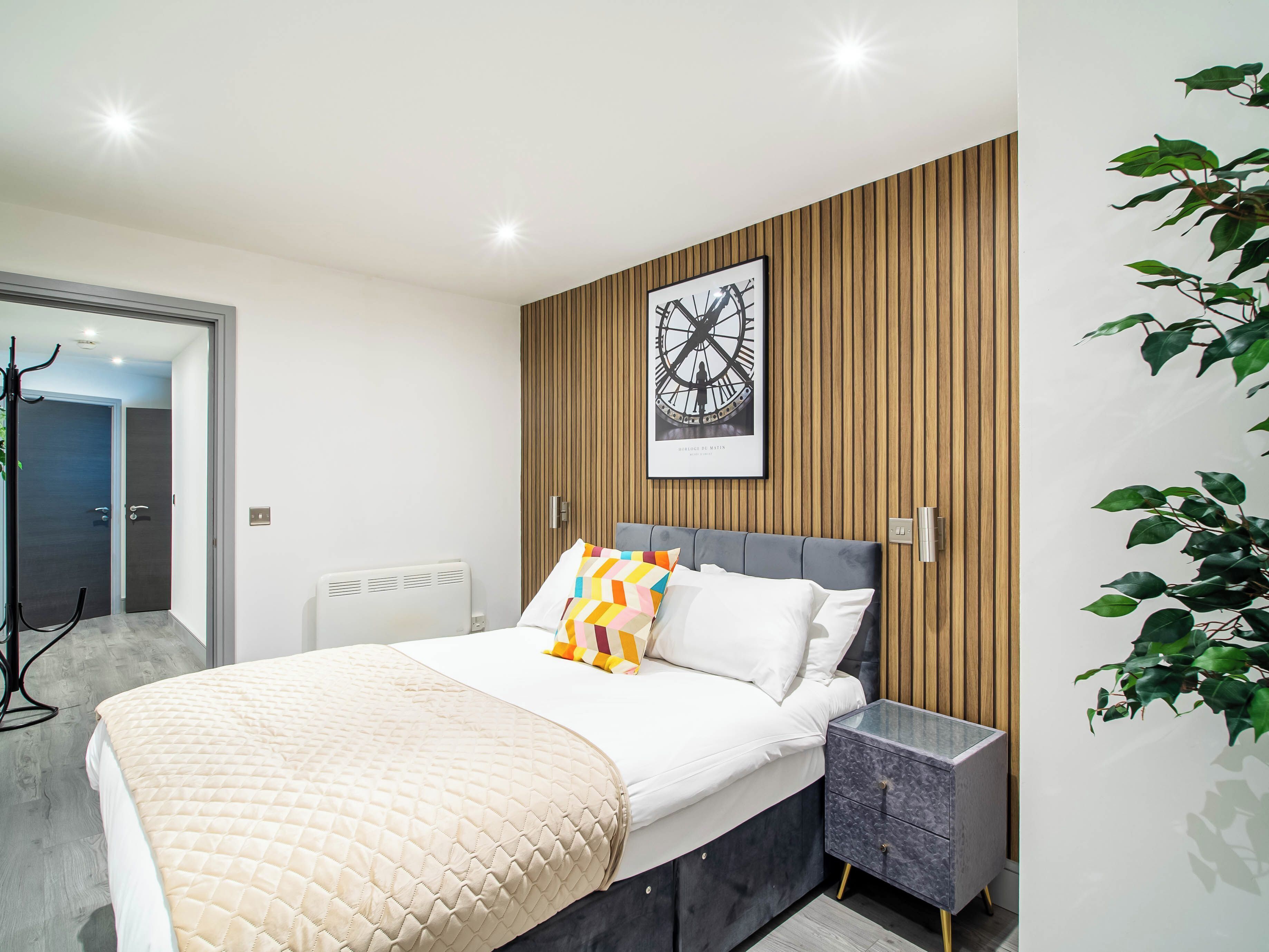 Quarto moderno com cama, mesa de cabeceira, radiador, painel de parede de madeira e uma moldura com imagem.