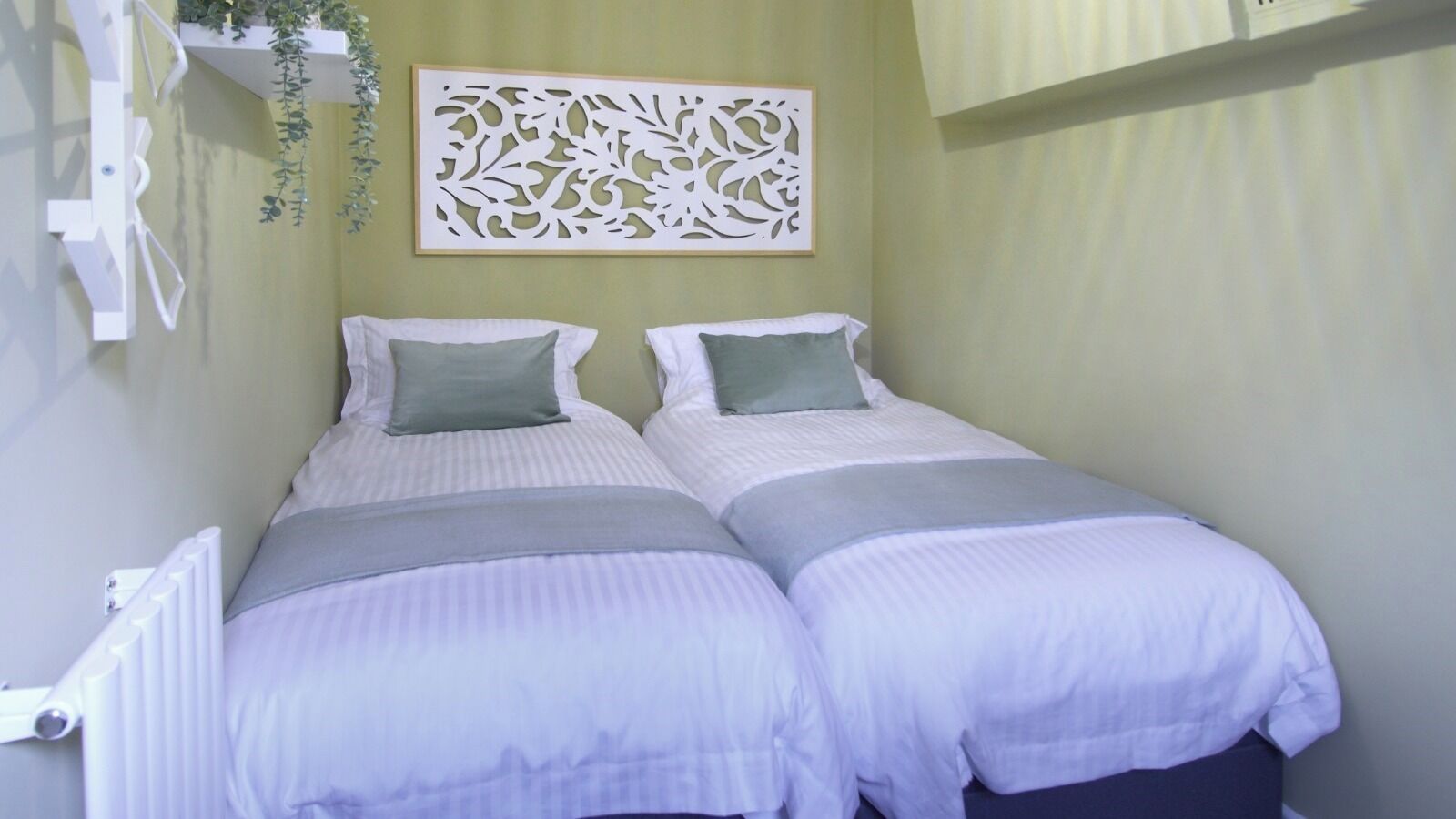 Quarto duplo com duas camas, um radiador, um painel decorativo na parede e uma planta pendente.