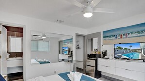 Quarto de hotel moderno com cama, televisão, cozinha compacta e uma janela grande com vista para a praia.