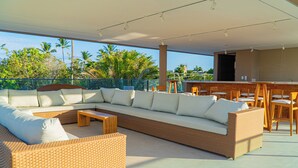 Uma área de lounge no terraço com um amplo sofá de vime, cadeiras de madeira e um bar.