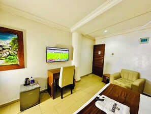 Quarto de hotel com televisão exibindo um evento esportivo, uma escrivaninha de madeira, uma cadeira, um sofá e um pequeno armário.