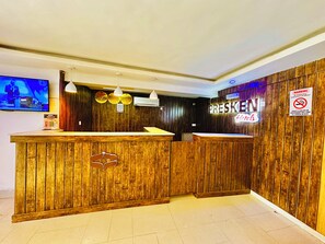 Uma mesa de recepção com painéis de madeira, uma televisão fixada na parede e uma placa com a inscrição 'PRESKEN Hotel'.