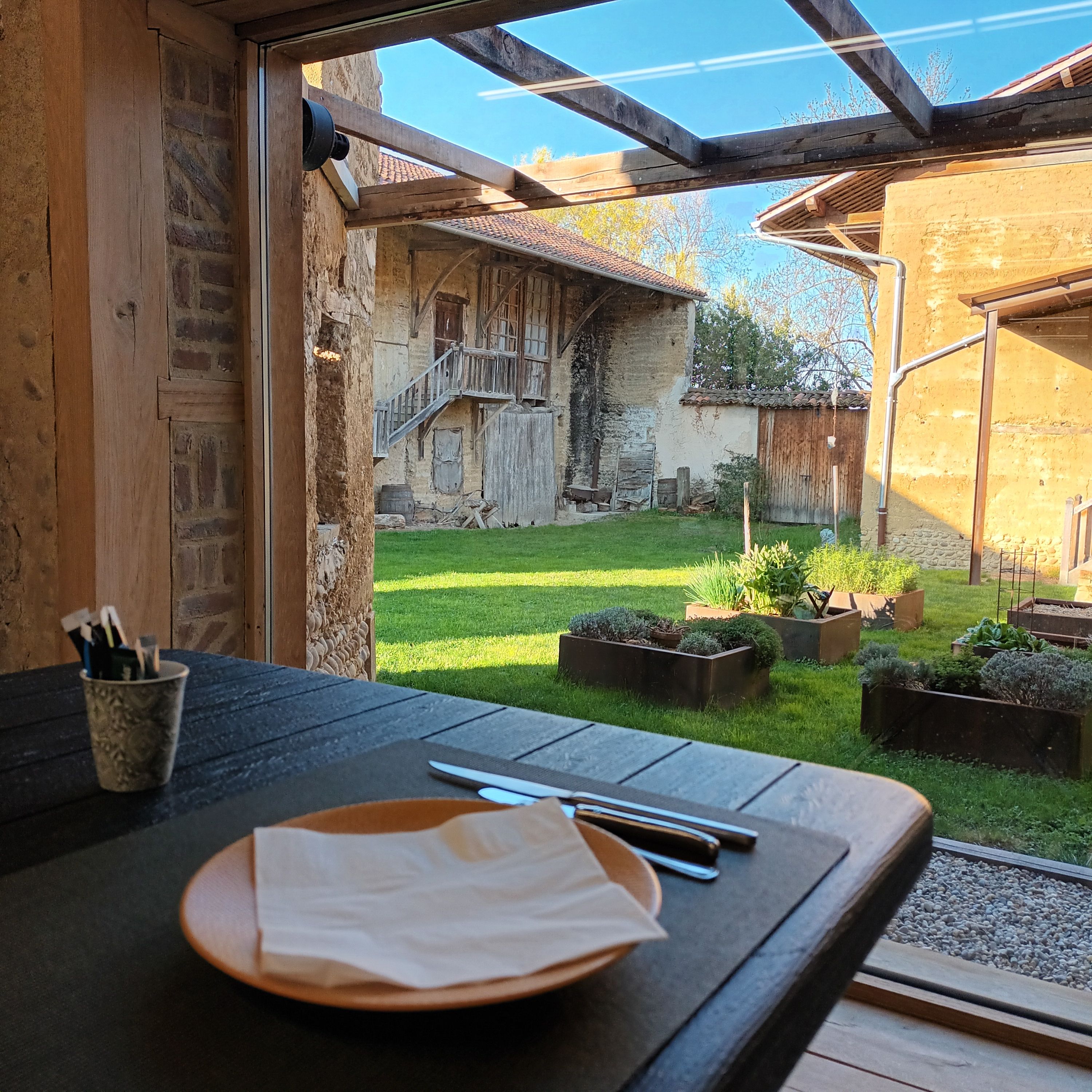 Uma mesa de madeira rústica com um prato e talheres, com vista para um jardim bem cuidado, com diversas plantas e um edifício de pedra tradicional ao fundo.