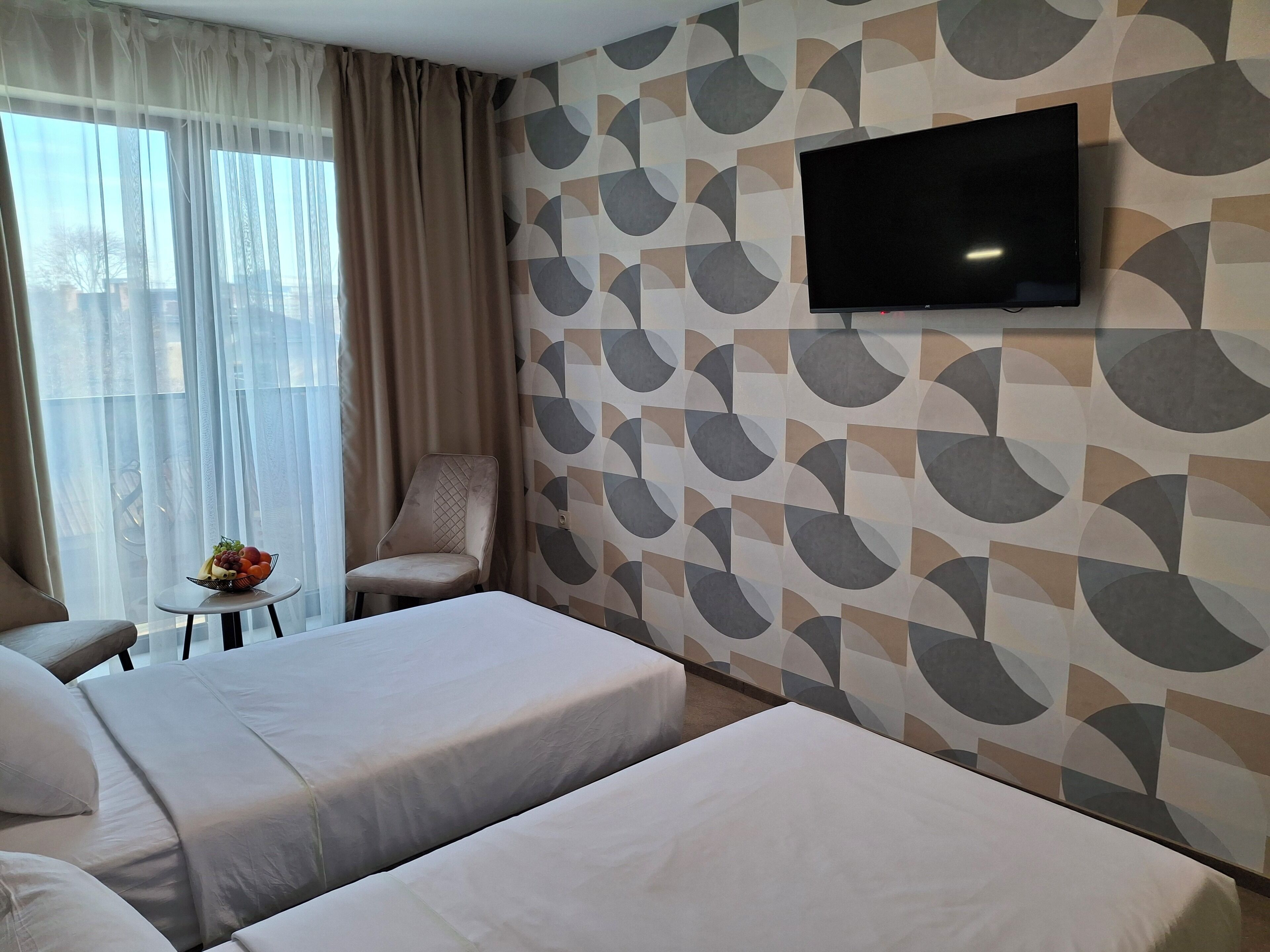 Quarto de hotel com duas camas, televisão na parede, uma cadeira, uma mesinha com uma fruteira e uma janela com cortinas.
