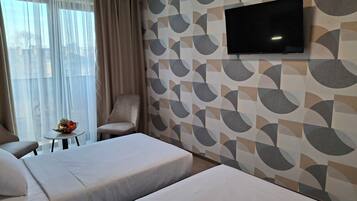 Quarto de hotel com duas camas, televisão na parede, uma cadeira, uma mesinha com uma fruteira e uma janela com cortinas.