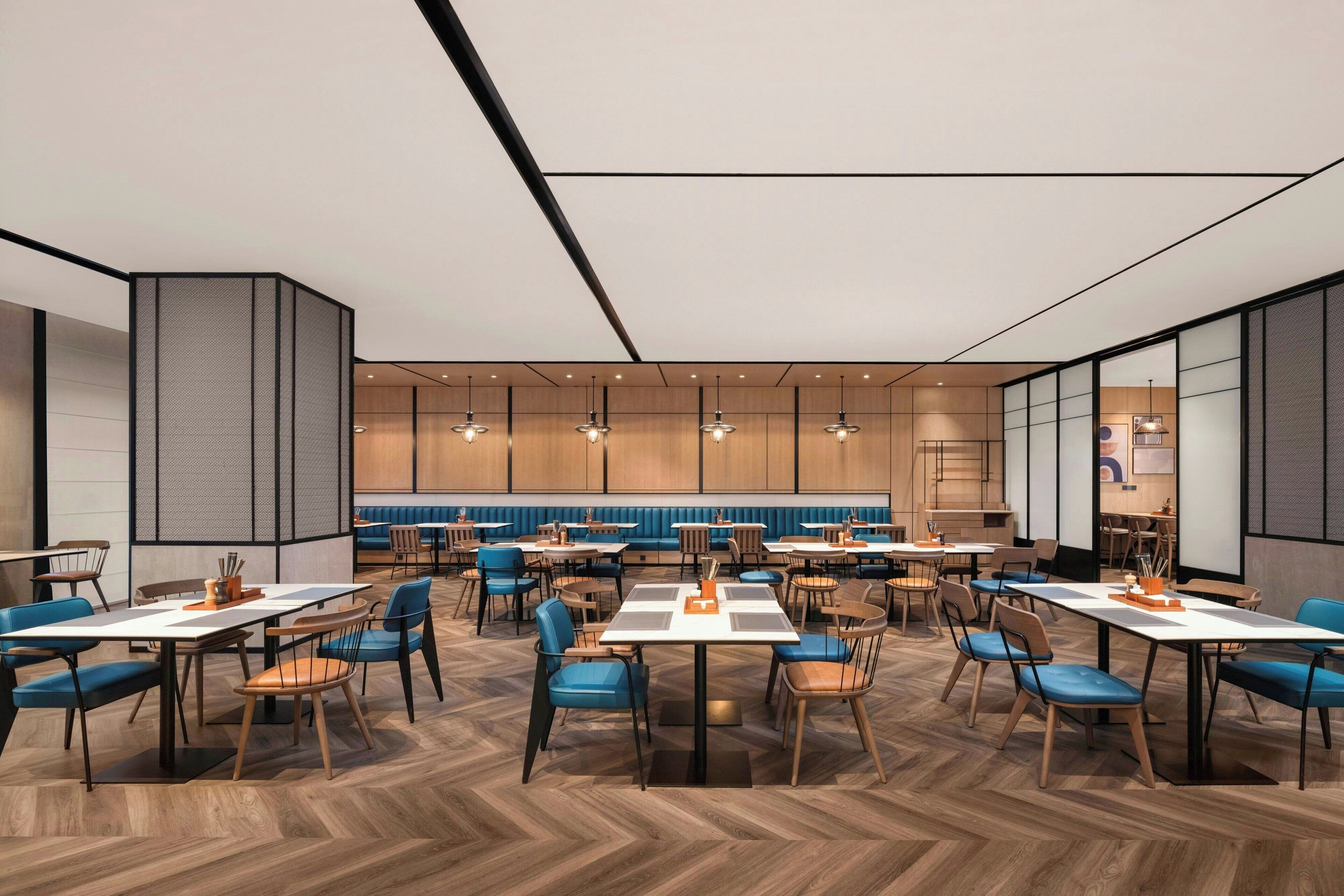Uma sala de jantar moderna com várias mesas e cadeiras, um piso de madeira em padrão losango e uma área de estar central com um banco azul.