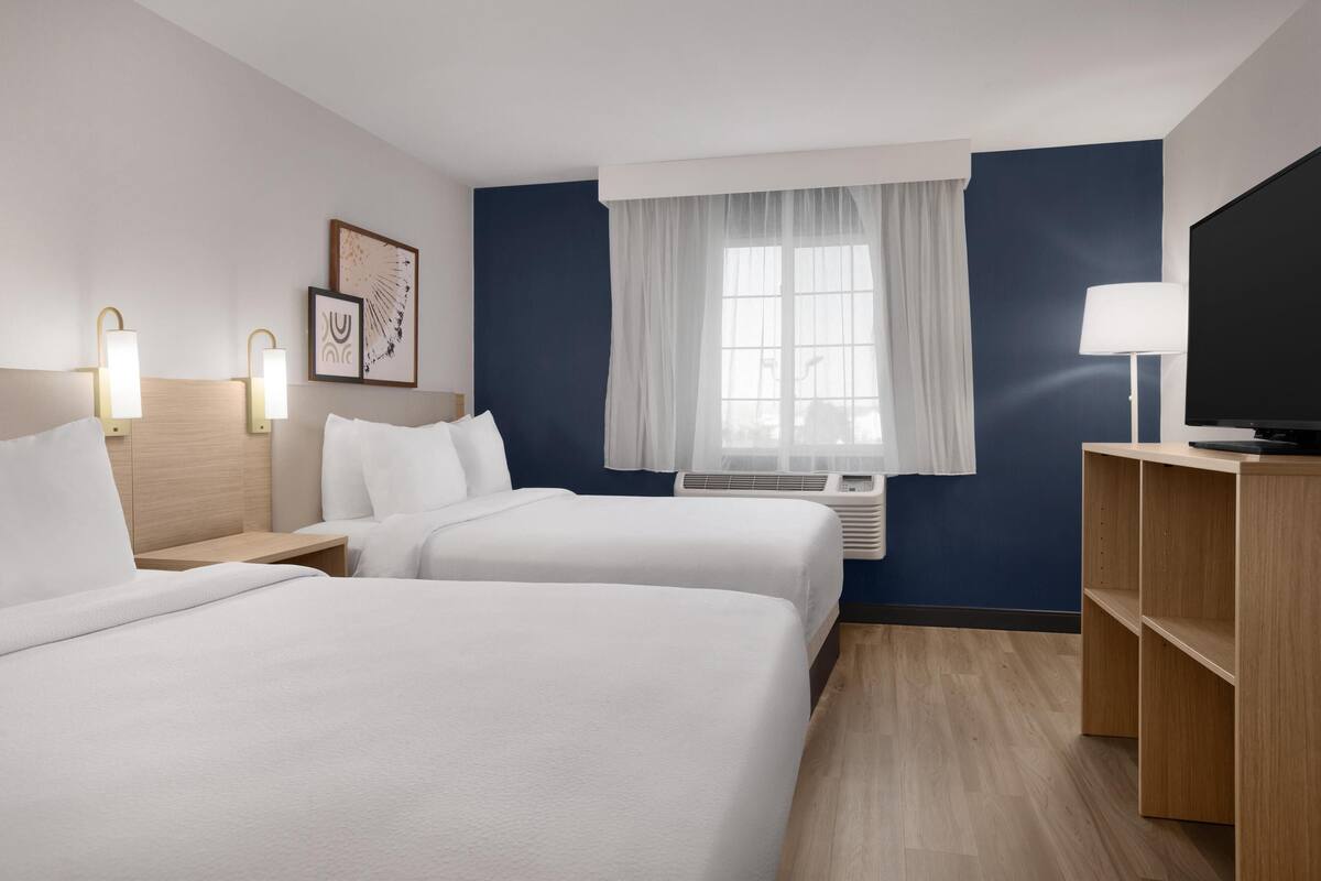 Quarto de hotel com duas camas, televisão de tela plana, escrivaninha e janela com cortinas leves.