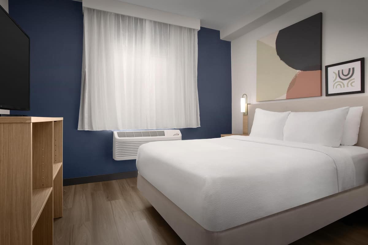Quarto de hotel com cama, televisão, escrivaninha e janela com cortinas.
