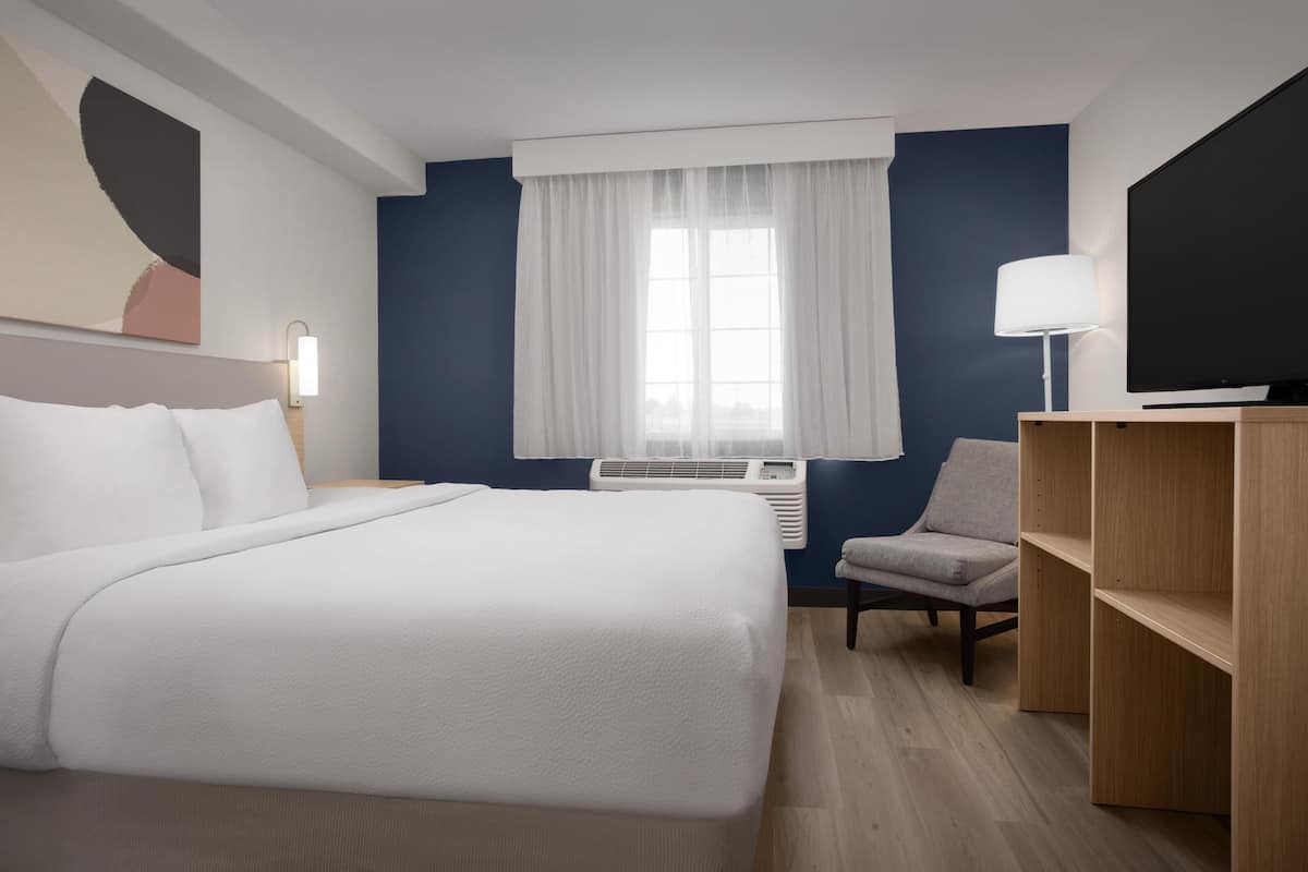 Quarto de hotel com uma cama grande, uma cadeira, uma televisão e uma janela com cortinas.