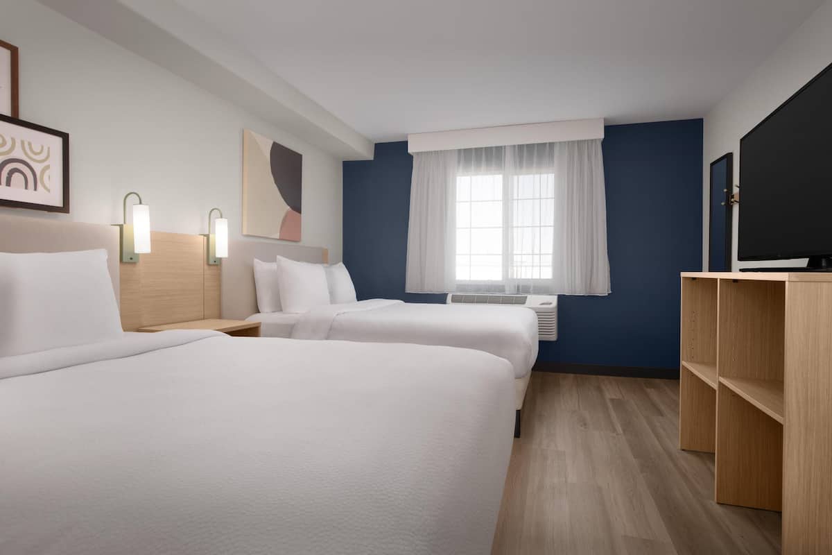 Quarto de hotel com duas camas, televisão, janela com cortinas e uma obra de arte na parede.