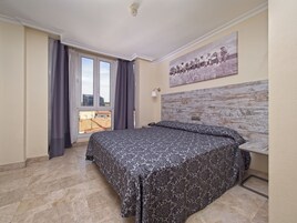 Quarto de hotel com cama, cabeceira, janela com cortinas, luminária de parede e pintura de paisagem urbana.