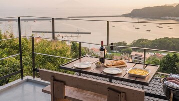 Varanda com mesa posta para refeição, com vista para a água e uma linha de costa distante.
