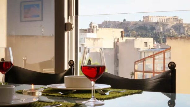Mesa de jantar com um copo de vinho tinto, uma vela e um prato de comida, com vista para uma paisagem urbana com arquitetura antiga.