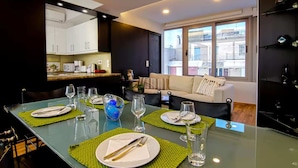 Uma sala de jantar moderna com mesa de vidro posta para quatro pessoas, um sofá ao fundo e uma cozinha visível por uma porta aberta.