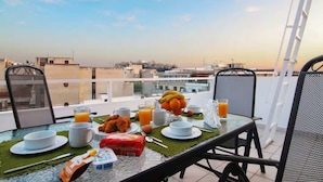 Mesa de café da manhã com pratos, xícaras e uma tigela em uma varanda com vista para a cidade.