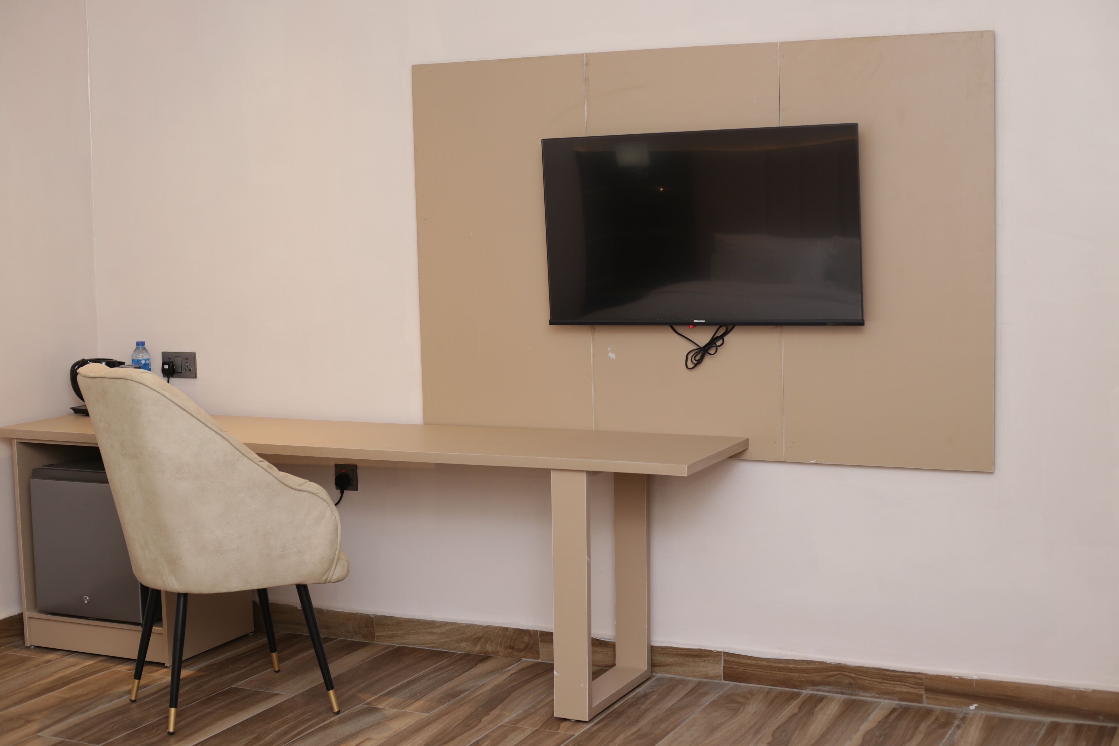 Quarto com televisão de tela plana presa a um painel bege, escrivaninha de madeira, cadeira bege e um pequeno armário.