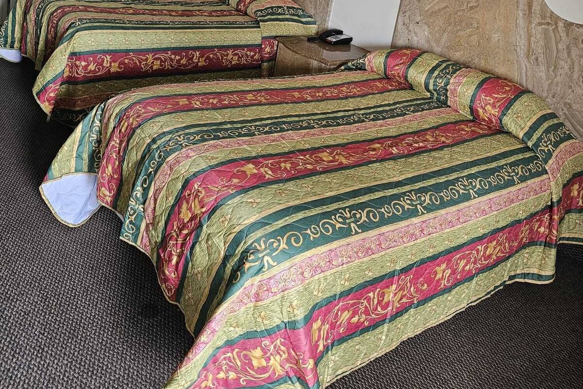 Quarto de hotel com duas camas, cada uma com cabeceira de madeira e colcha estampada. Há duas mesas de cabeceira com luminárias, e uma pequena estante com objetos decorativos na parede.