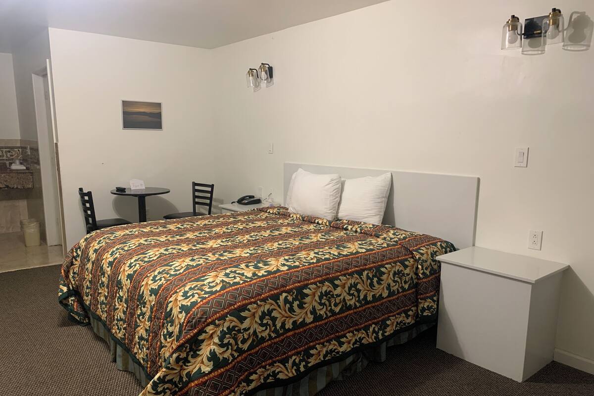 Quarto bem arrumado com colcha estampada, mesa de cabeceira, cadeira, pequena mesa com telefone e luminária fixada na parede.