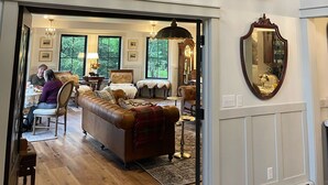 Uma sala de estar ampla com um grande sofá de couro marrom, um espaço de jantar com mesa e cadeiras, e vista para uma paisagem verde exuberante através de grandes janelas.