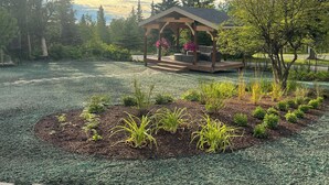 Um gazebo com telhado de madeira e bancos, cercado por um jardim com diversas plantas e um caminho de cascalho com lajes de pedra.