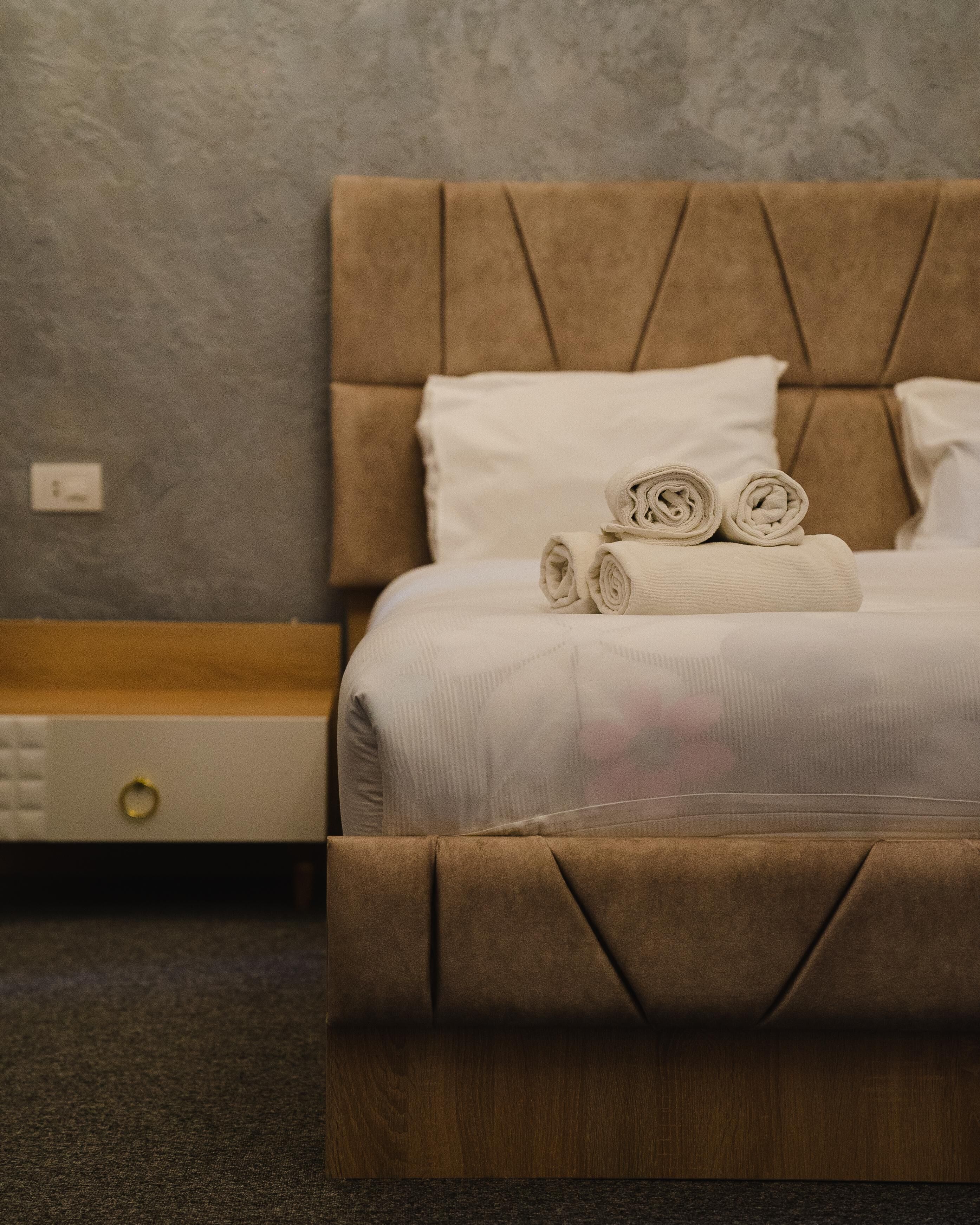 Cama bem arrumada com toalhas dobradas, mesa de cabeceira de madeira e parede texturizada cinza.