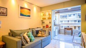 Sala de estar com sofá, almofadas, um quadro e cozinha visível através de uma porta aberta.