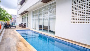 Uma piscina com borda azulejada, ao lado de um edifício com amplas janelas e fachada branca.