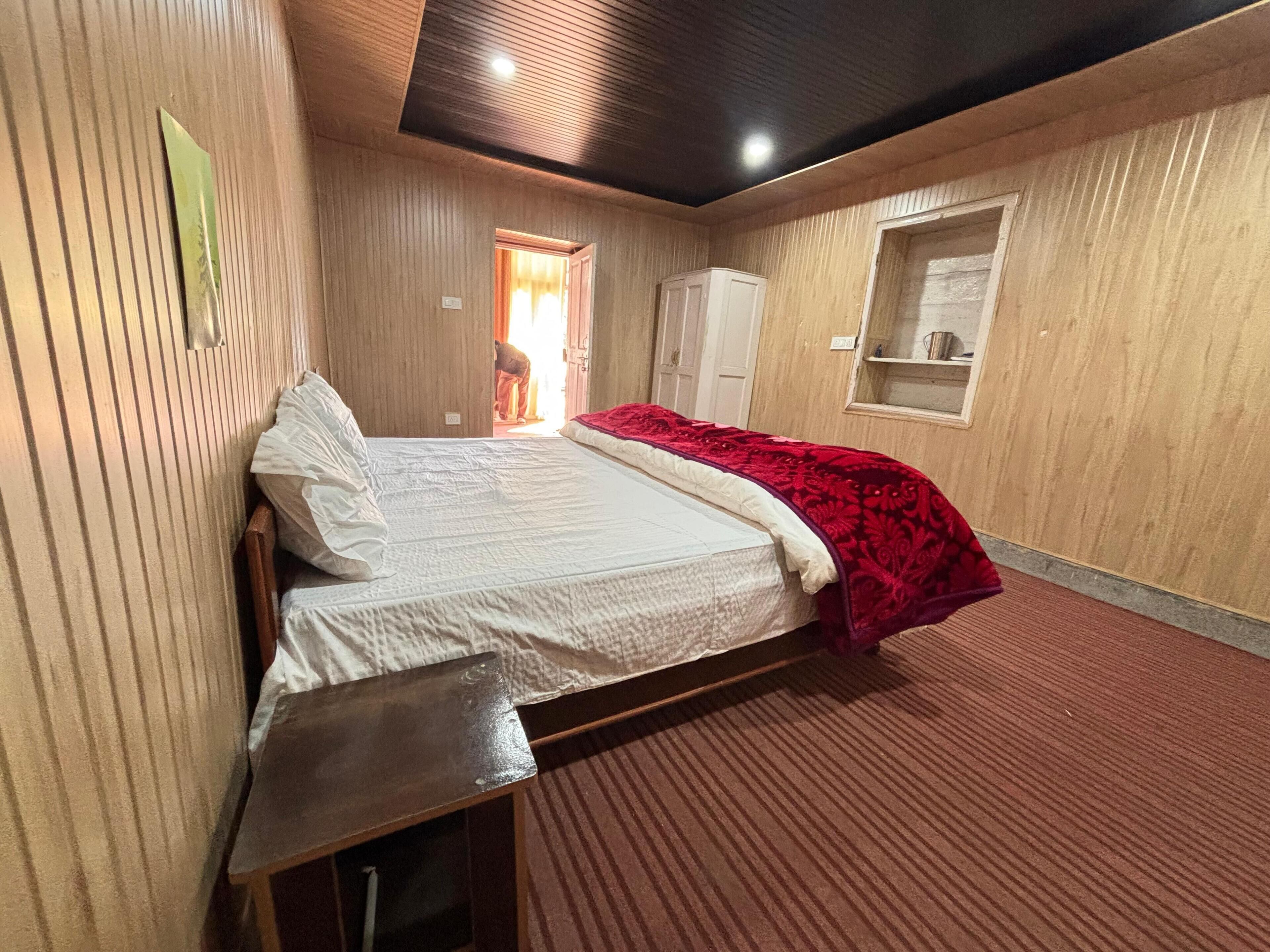 Quarto de hotel com uma cama, um guarda-roupa, uma pequena prateleira e uma porta que dá para outro aposento.