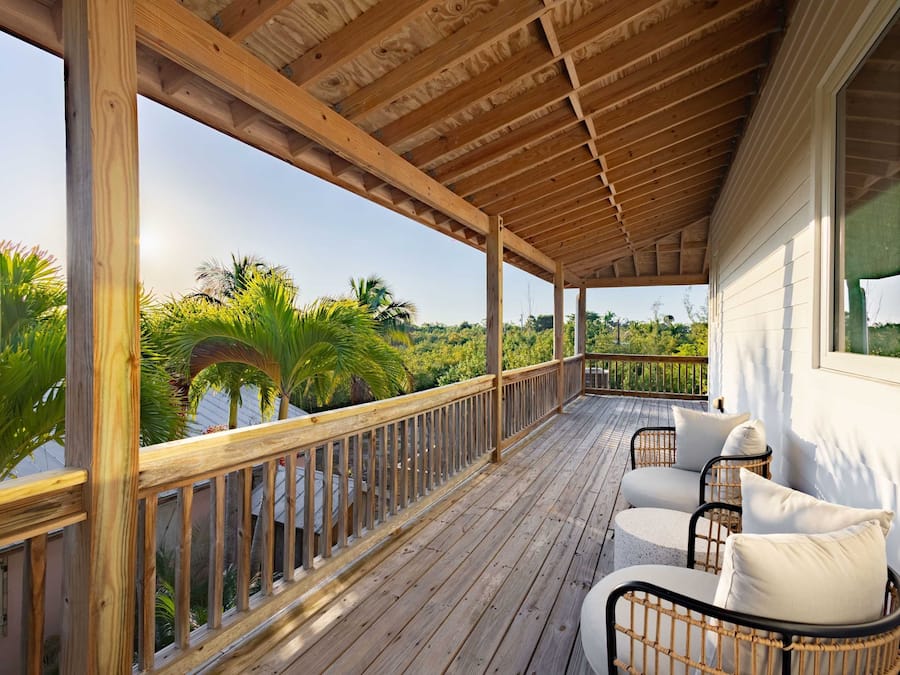Um deck de madeira com guarda-corpo, cadeiras de vime e vista para o verde.