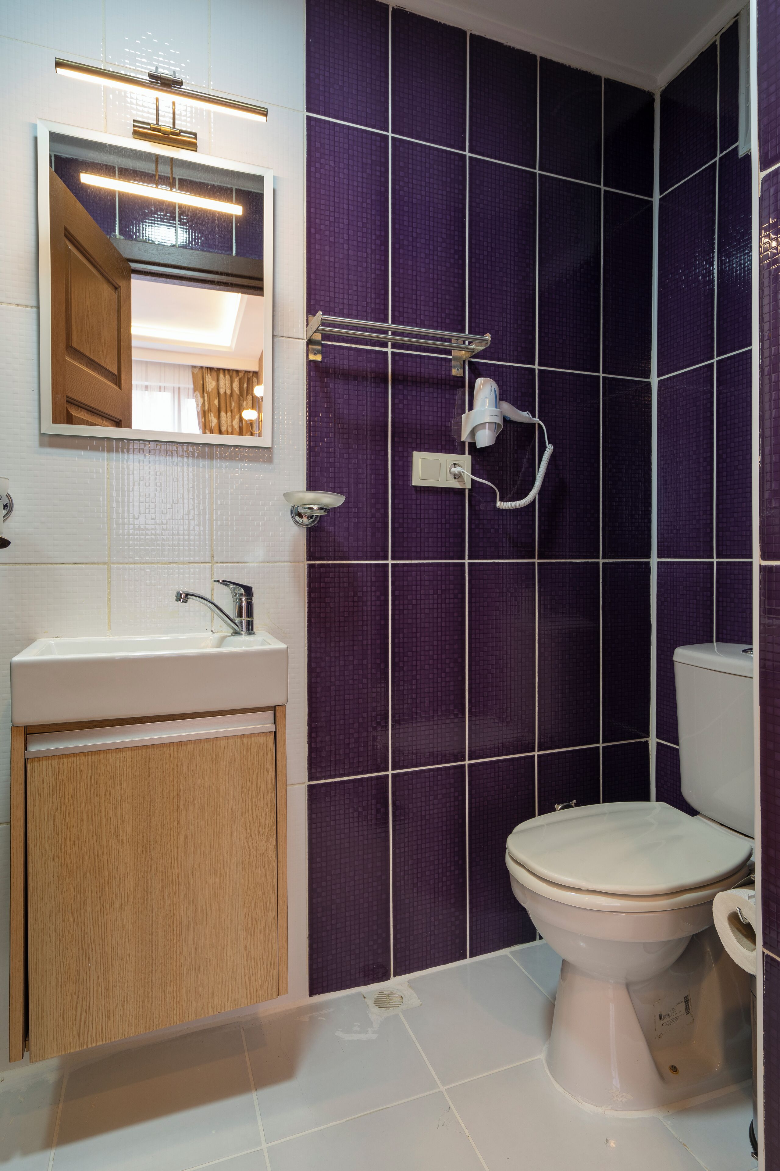 Um banheiro com pia branca, espelho e vaso sanitário, e paredes revestidas com azulejos roxos.