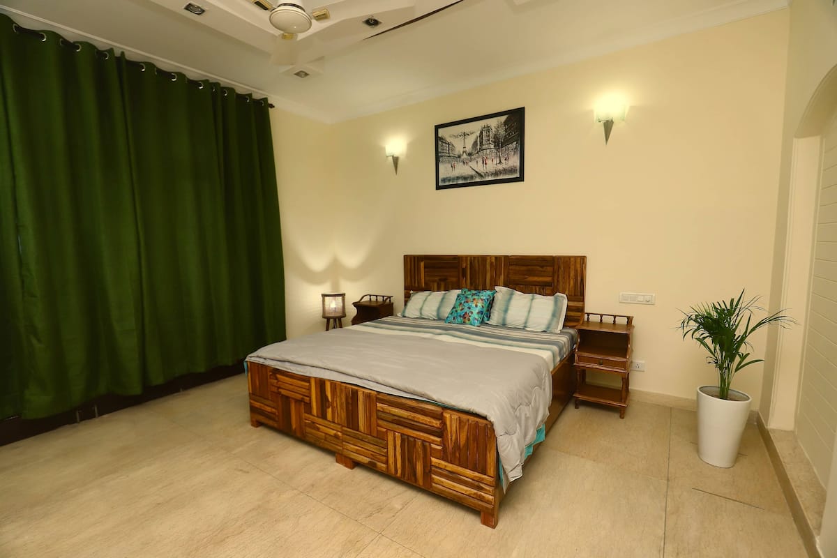 Quarto com cama de madeira, cortinas verdes, quadro na parede, abajures e uma planta em vaso.
