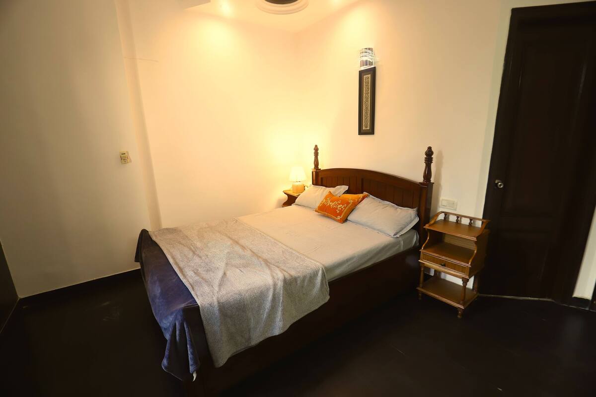 Uma cama bem arrumada com cabeceira de madeira, uma mesa de cabeceira com abajur e um criado-mudo de madeira.