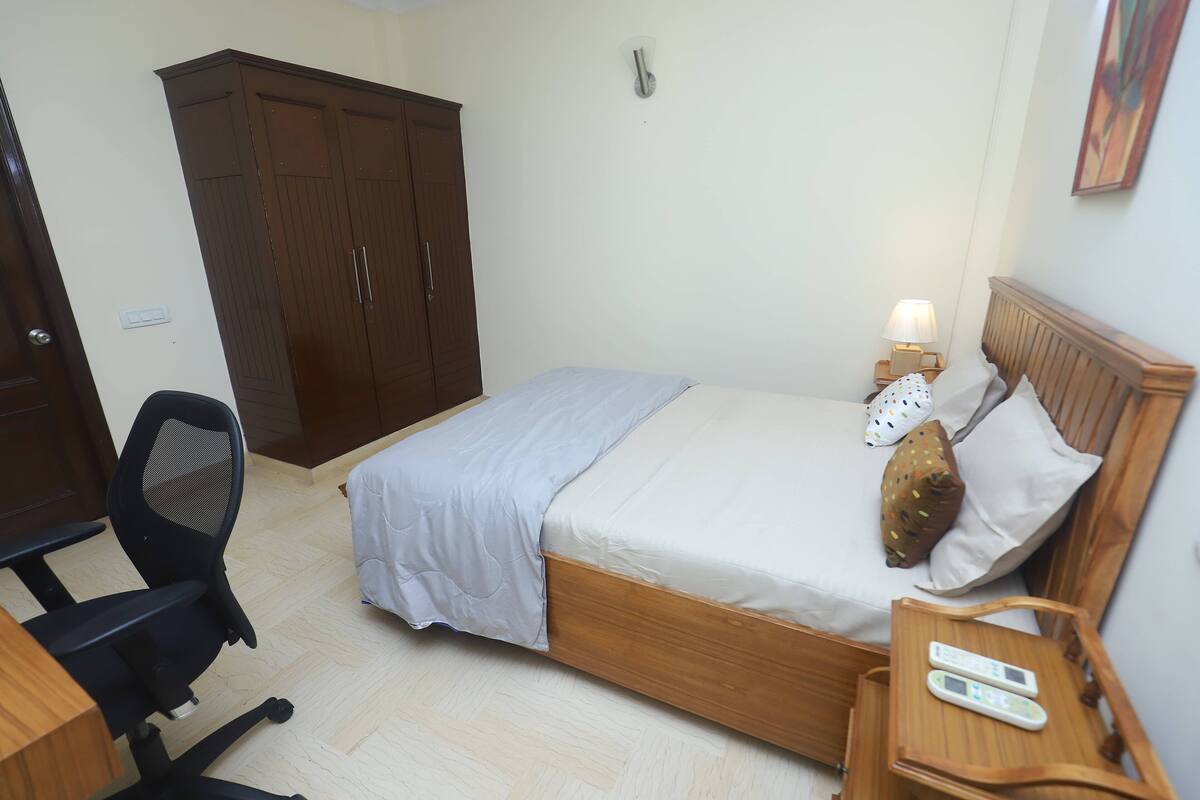 Quarto com cama, mesa de cabeceira, abajur, cadeira, escrivaninha, guarda-roupa e relógio de parede.
