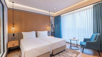 Quarto de hotel moderno com duas camas, uma mesa redonda pequena, uma poltrona azul e uma janela grande com cortinas.