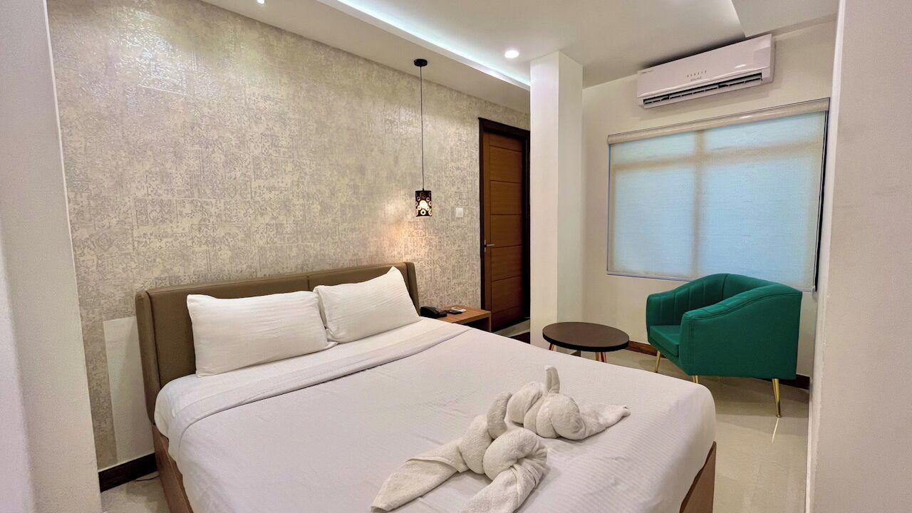 Quarto de hotel com uma cama grande, uma mesa redonda pequena, uma cadeira verde e uma janela com persianas.