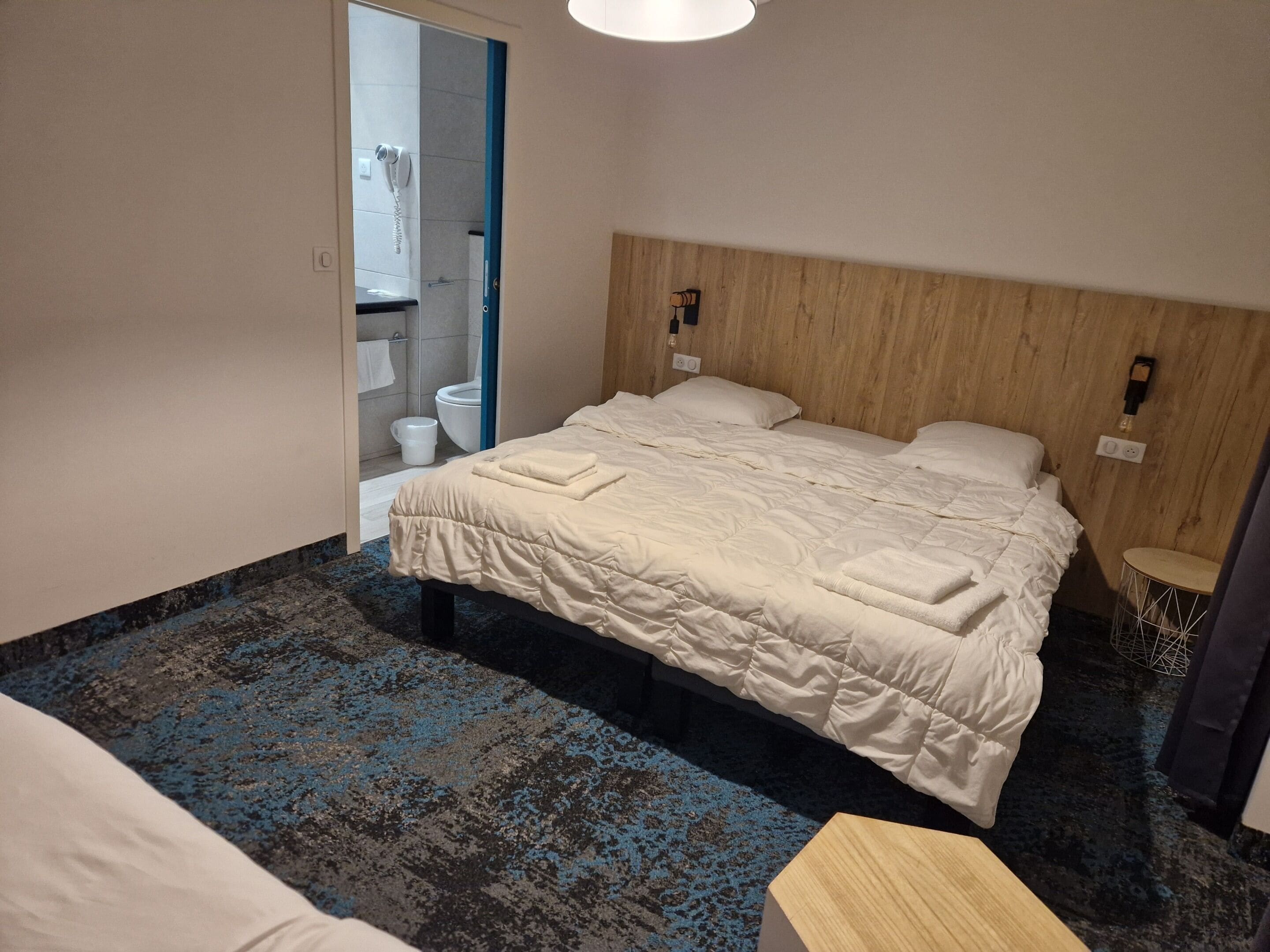 Quarto de hotel com cama, cabeceira de madeira, pequena mesa de apoio lateral e banheiro visível por uma porta aberta.