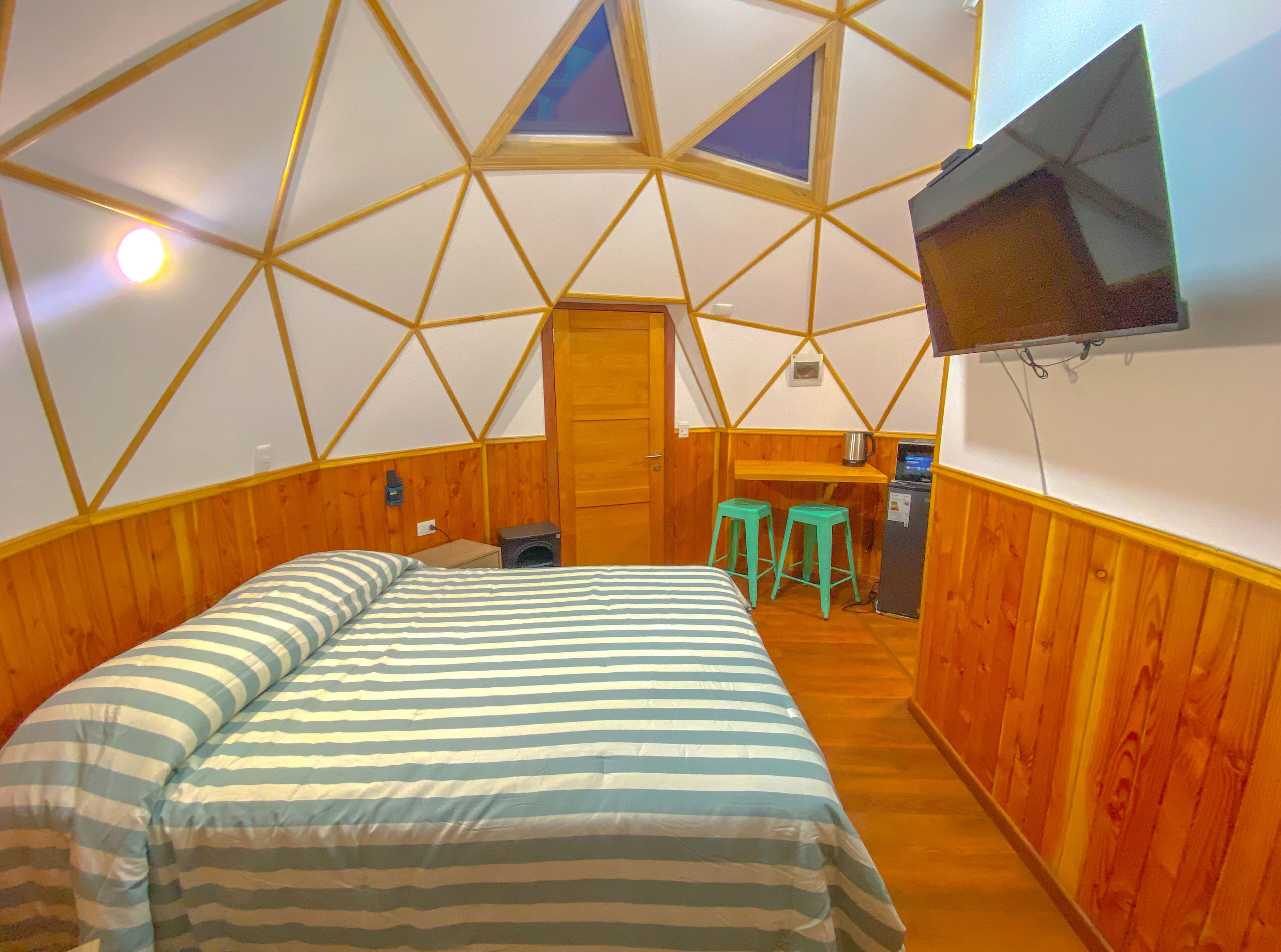 Quarto com uma cama, dois banquinhos, uma televisão e uma cozinha compacta.