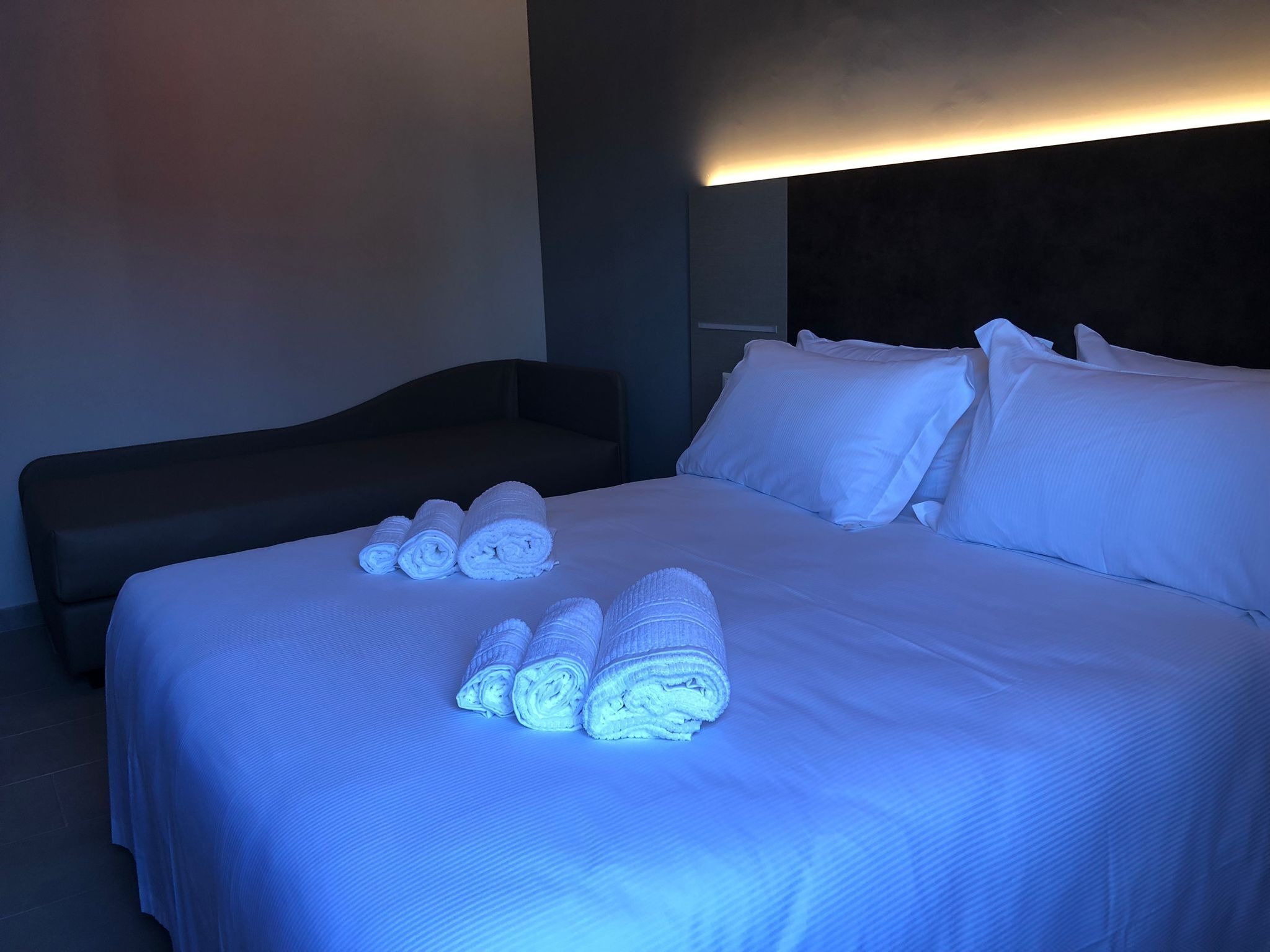 Cama bem arrumada com roupa de cama branca e toalhas dobradas, cabeceira escura e um banco à esquerda.