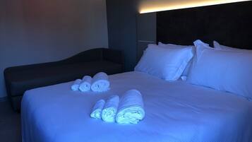 Cama bem arrumada com roupa de cama branca e toalhas dobradas, cabeceira escura e um banco à esquerda.