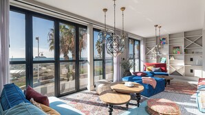 Uma sala de estar ampla com janelas grandes, um sofá azul, uma mesa de centro de madeira e uma estante de livros.