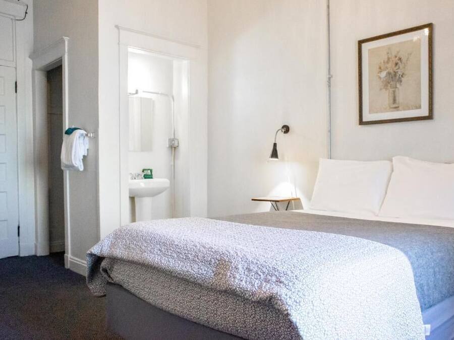 Quarto de hotel com uma cama, uma mesa de cabeceira com abajur, um quadro na parede e um banheiro visível por uma porta aberta.