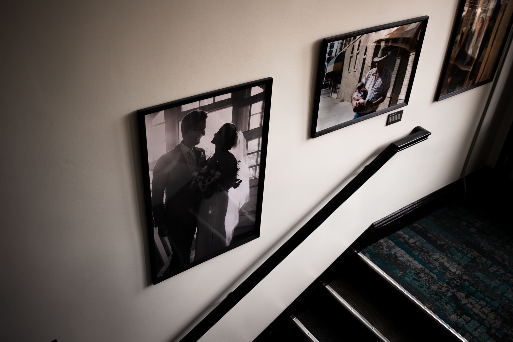 Um corredor com fotografias emolduradas na parede, uma escada com carpete estampado e um candeeiro fixado na parede.
