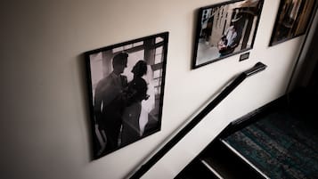 Um corredor com fotografias emolduradas na parede, uma escada com carpete estampado e um candeeiro fixado na parede.