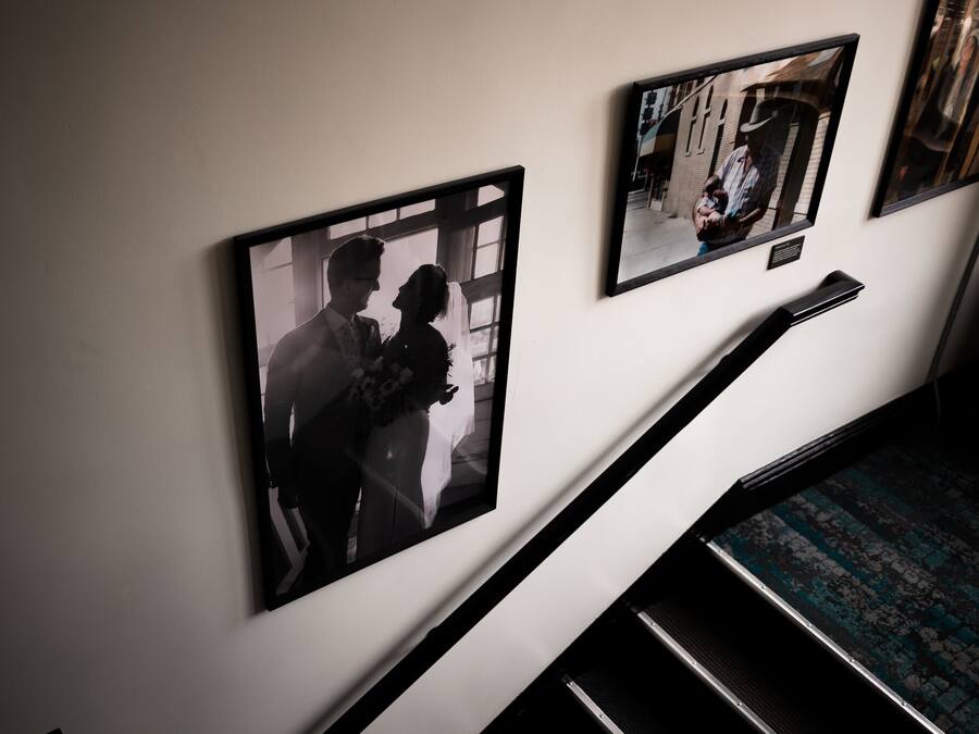 Um corredor com fotografias emolduradas na parede, uma escada com carpete estampado e um candeeiro fixado na parede.