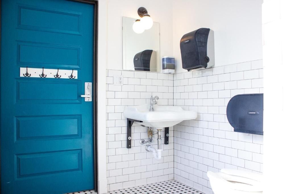 Um banheiro com porta azul, paredes com azulejos brancos, pia, espelho e dosador de sabão.