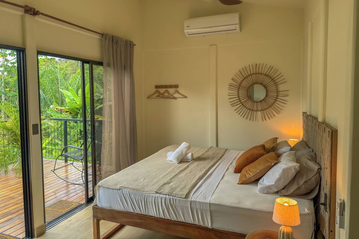 Quarto com cama, ventilador de teto, unidade de ar condicionado, espelho fixado na parede e vista para o verde através de uma porta de correr.