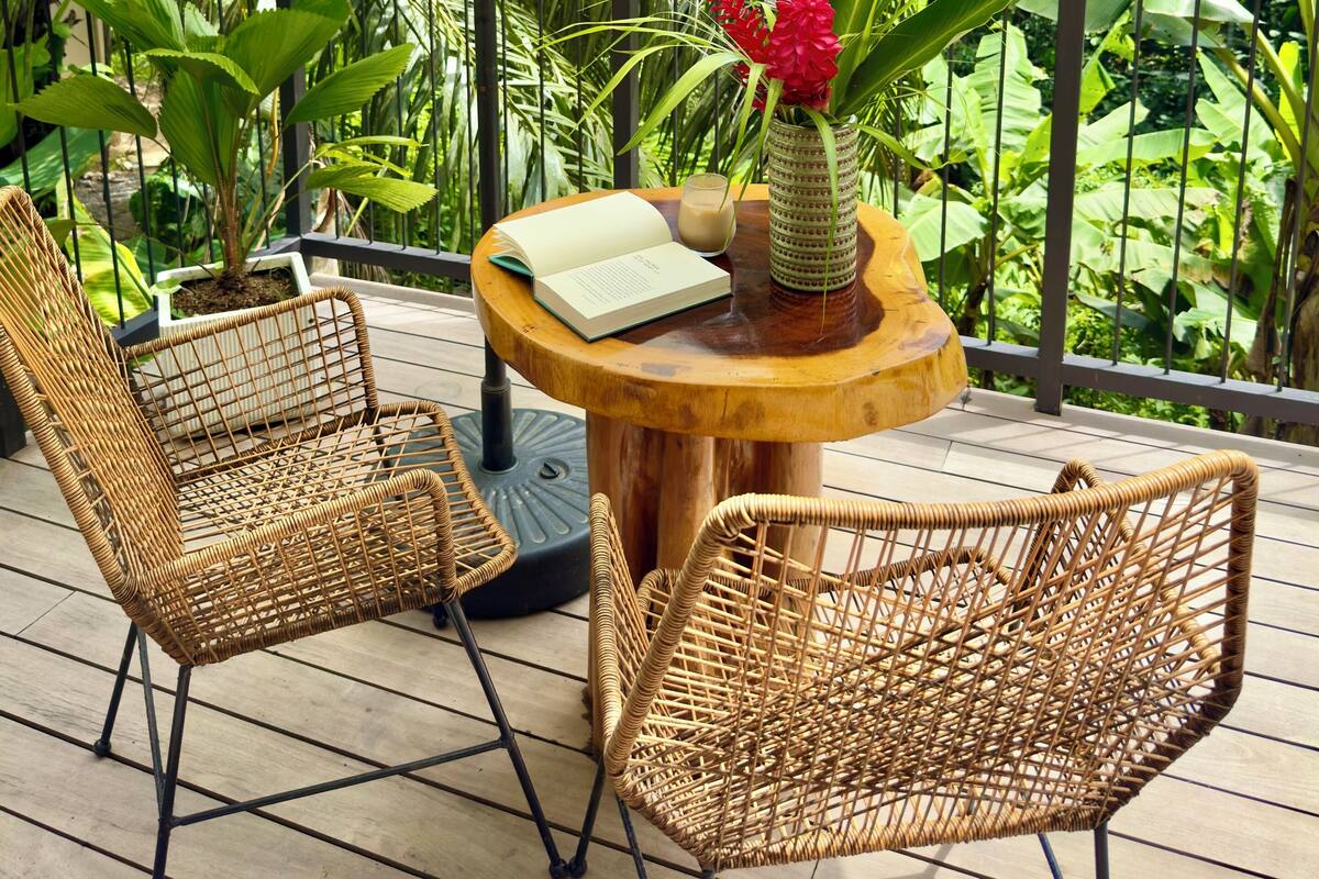 Uma área de estar coberta ao ar livre, com cadeiras de vime e uma mesa, rodeada por plantas tropicais exuberantes e um guarda-sol grande.