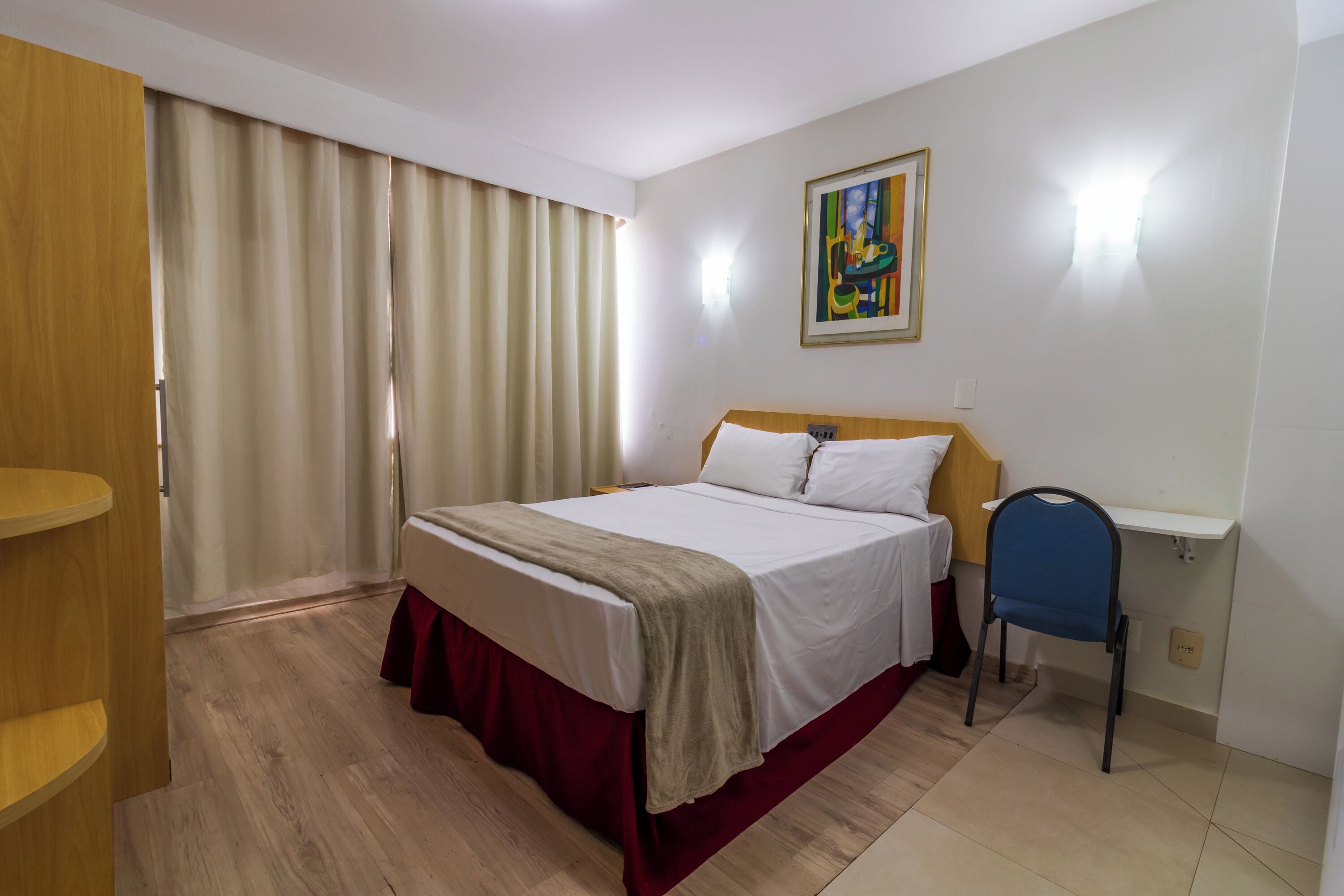 Quarto de hotel com cama, escrivaninha, cadeira, guarda-roupa e um quadro na parede.
