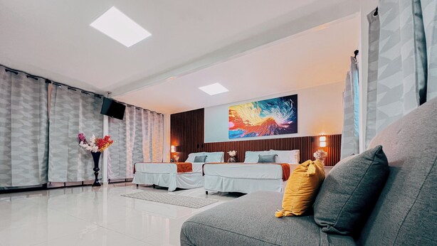 Quarto de hotel moderno com um amplo sofá cinzento, uma cama com roupa de cama branca, uma televisão de ecrã plano na parede e um quadro abstrato colorido sobre a cama.