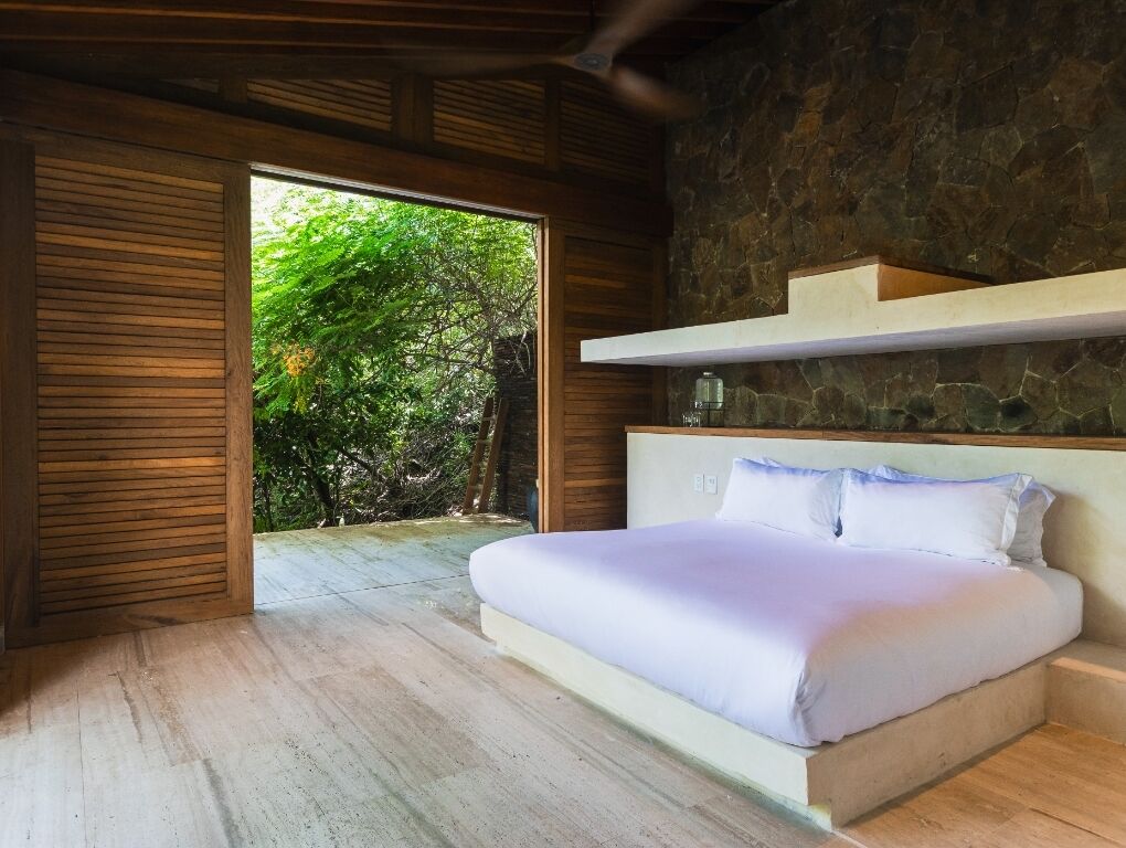 Quarto com uma cama grande, paredes de madeira, parede decorativa de pedra e vista para o verde lá fora.